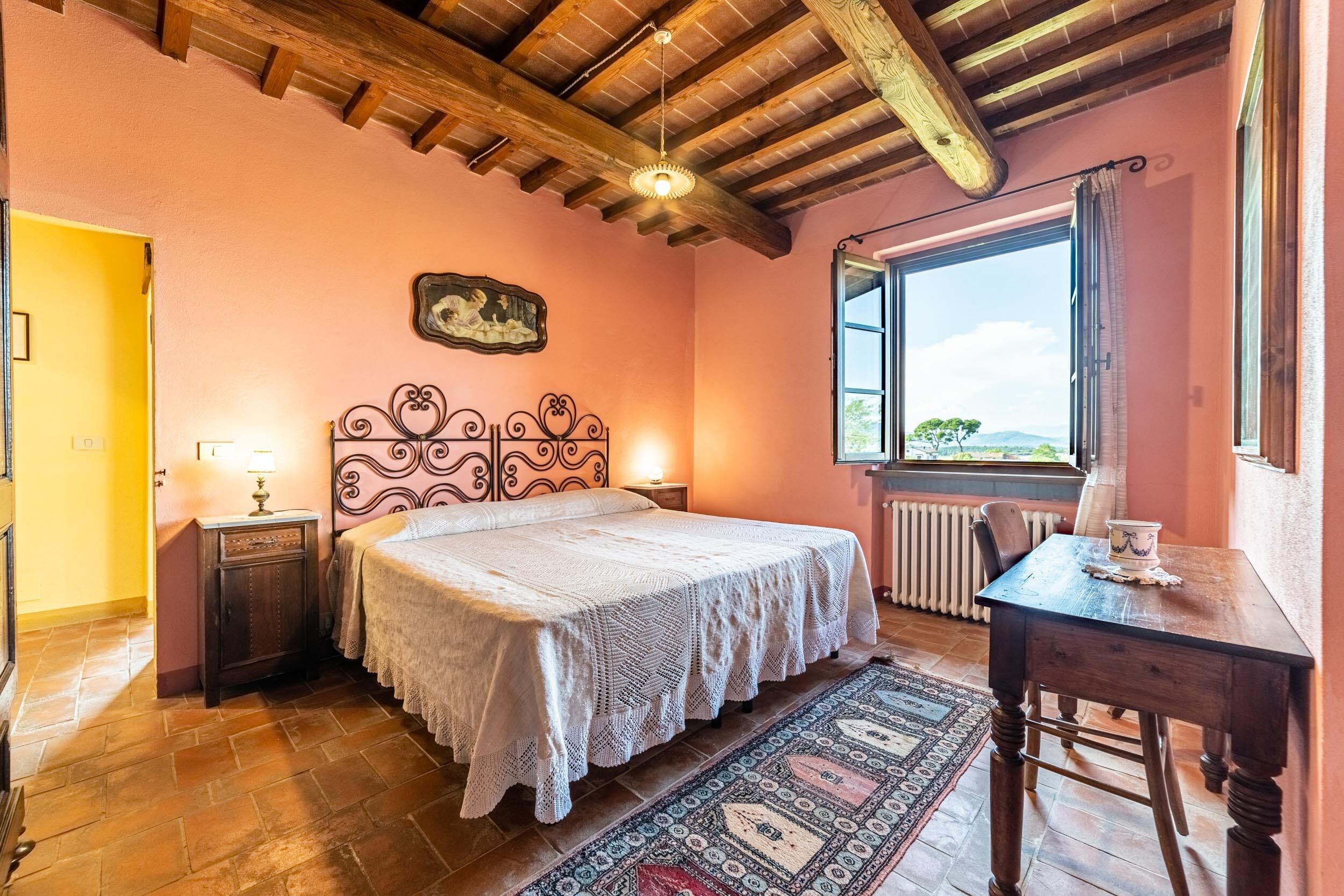 Quarto com teto de madeira, cama com colcha branca, mesa de cabeceira com abajur, janela com vista e mesa de madeira com uma xícara.