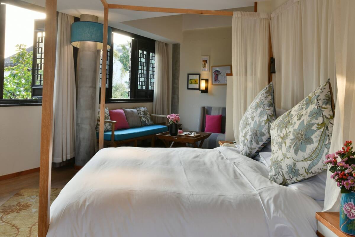 Quarto com cama de dossel, área de estar com sofá e poltronas, e janela com cortinas.