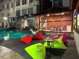 Uma área à beira da piscina com bar, pufes coloridos e uma parede de pedra.