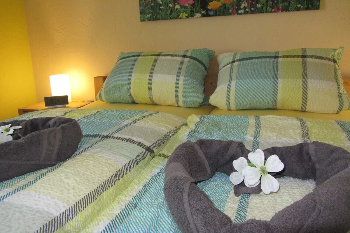 Uma cama com um edredom verde e branco quadriculado, duas almofadas com padrão semelhante e uma mesa de cabeceira com um relógio marcando 8:54.