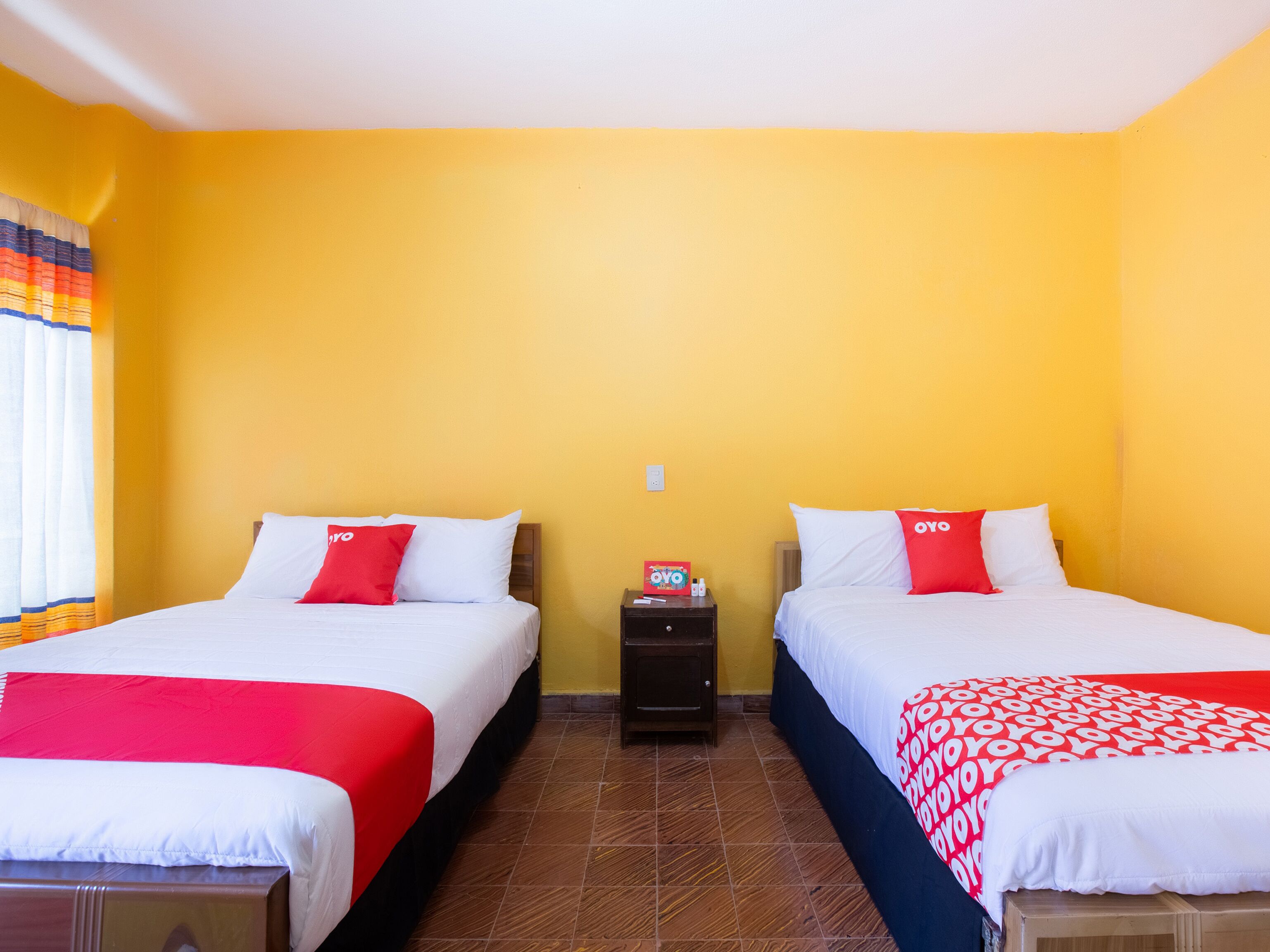 Quarto de hotel com duas camas, cada uma com colcha vermelha e branca, e uma mesinha de cabeceira pequena entre elas.