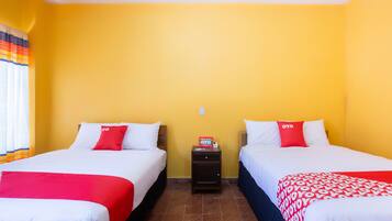 Quarto de hotel com duas camas, cada uma com colcha vermelha e branca, e uma mesinha de cabeceira pequena entre elas.