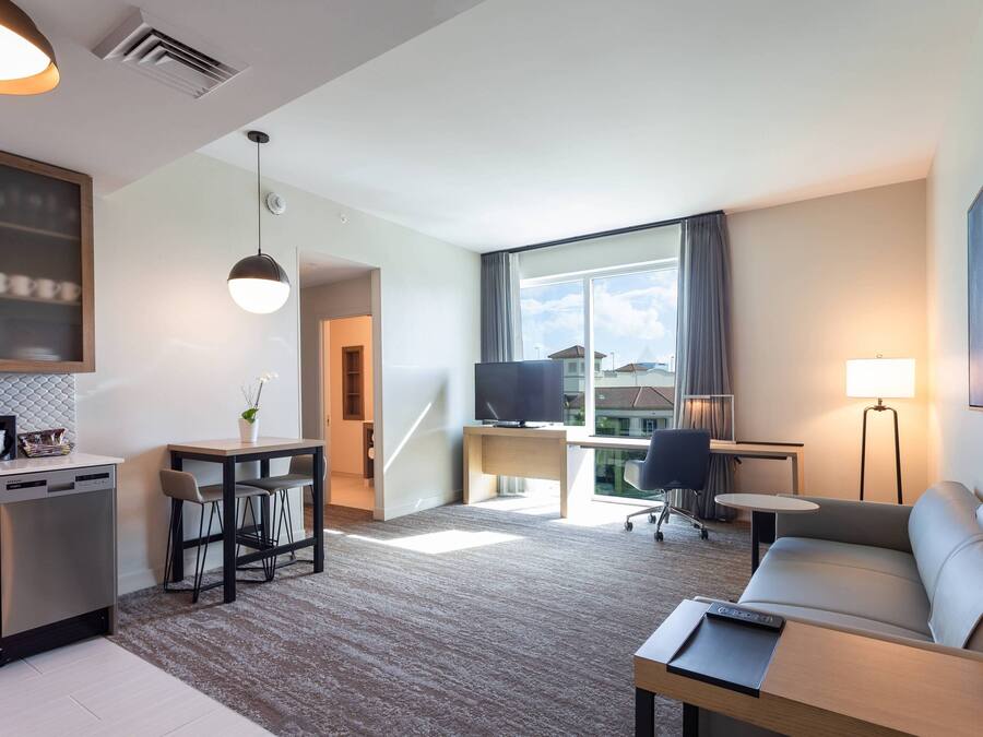 Quarto de hotel moderno com cozinha, mesa de jantar e cadeiras, sofá, escrivaninha com computador e uma grande janela com vista para a cidade.