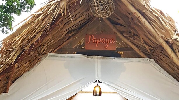 Uma construção com telhado de palha, com duas camas, uma luminária pendente e um letreiro com a palavra 'Papaya'.