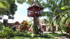 Uma estrutura de cabana na árvore com telhado de palha, rodeada por vegetação exuberante e um pequeno jardim.