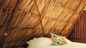 Um quarto com telhado de colmo, uma cama com lençol branco e travesseiro com padrão de abacaxi, uma pequena mesa de cabeceira de madeira com uma planta e um tapete estampado.