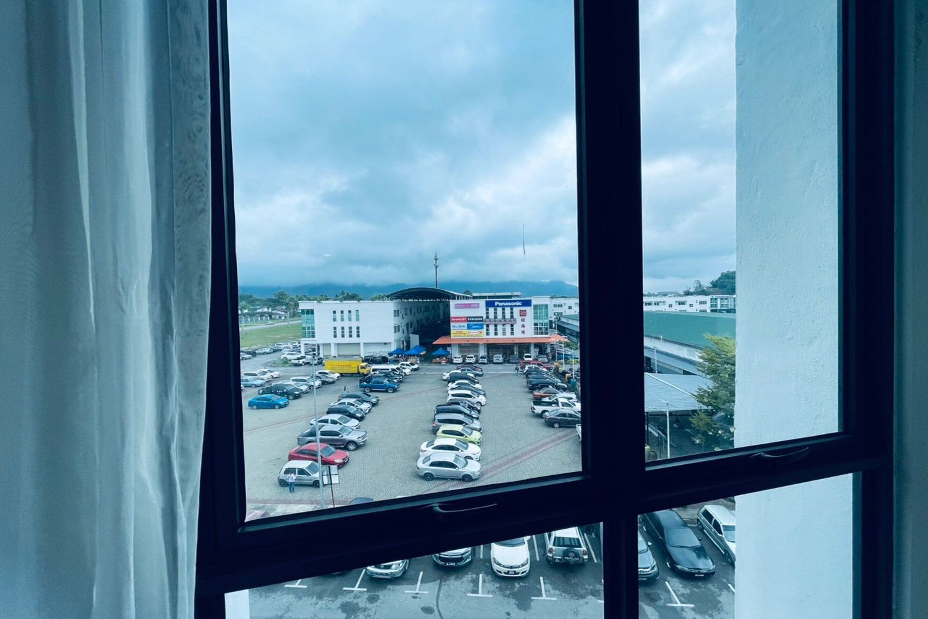 Vista da janela sobre um estacionamento com vários carros e um edifício com a placa 'Panasonic'.