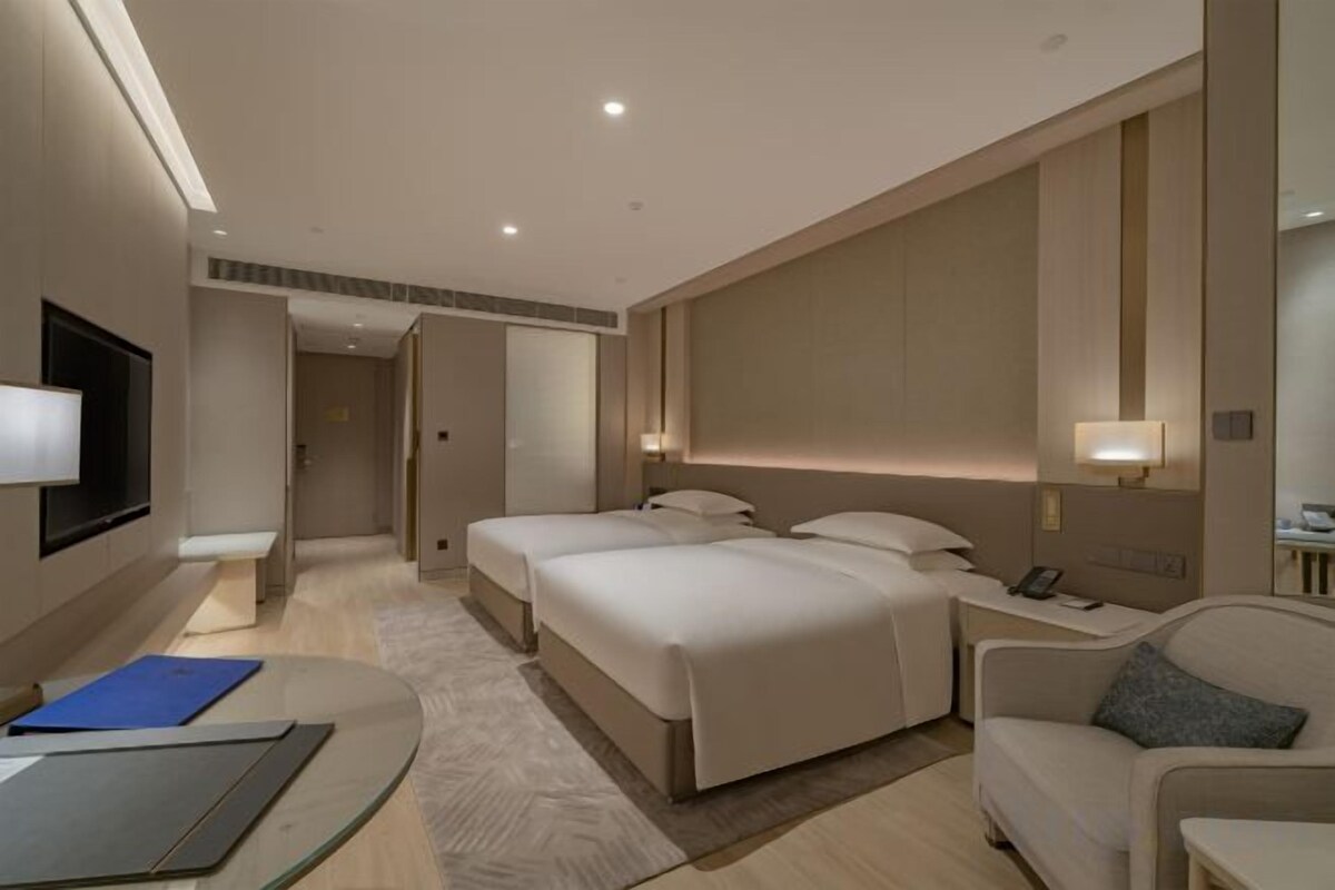 Quarto de hotel com duas camas, uma escrivaninha, uma cadeira, uma televisão e banheiro.