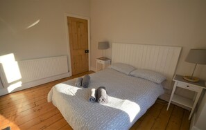 Quarto com piso de madeira, cama com colcha estampada, duas toalhas enroladas na cama, porta de madeira, radiador branco e duas mesas de cabeceira com luminárias.