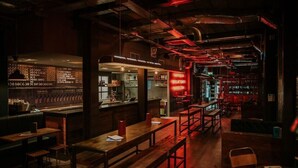 Um bar com iluminação baixa, mesas e bancos de madeira, canos aparentes e um letreiro neon.