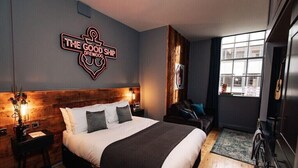 Quarto de hotel moderno com uma cama grande, cabeceira de madeira, um letreiro neon com a frase "The Good Ship Brewdog", um pequeno espaço de estar com uma cadeira e uma mesa, e uma janela com vista para a cidade.