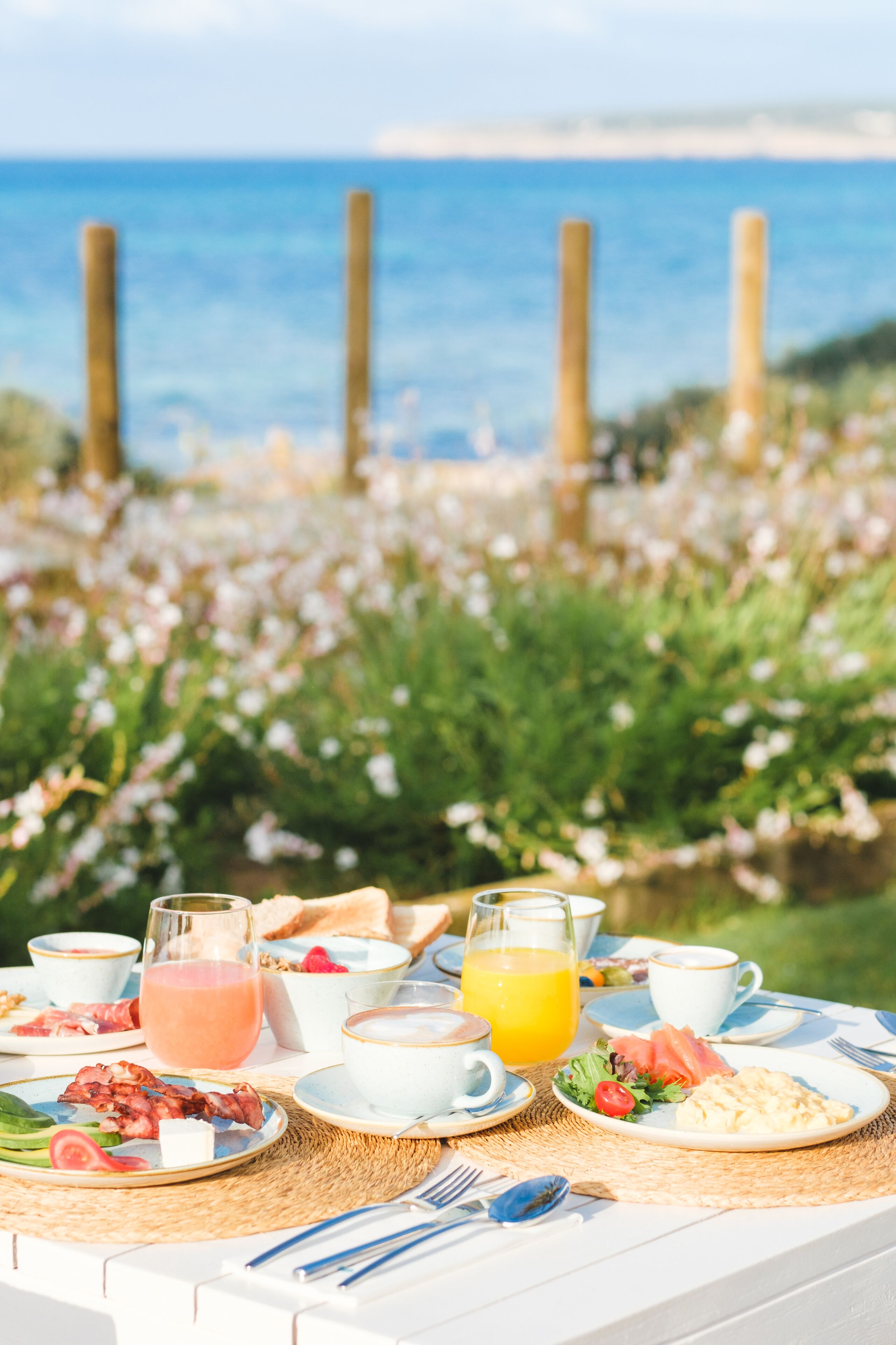 Mesa de café da manhã com pratos de comida, xícaras e copos, com o cenário de uma praia e flores ao fundo.