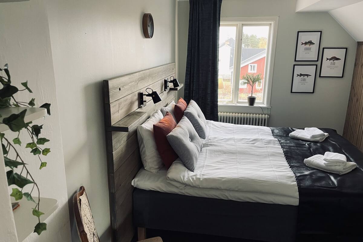 Cama bem arrumada com almofadas, mesa de cabeceira com abajur e janela com cortinas.