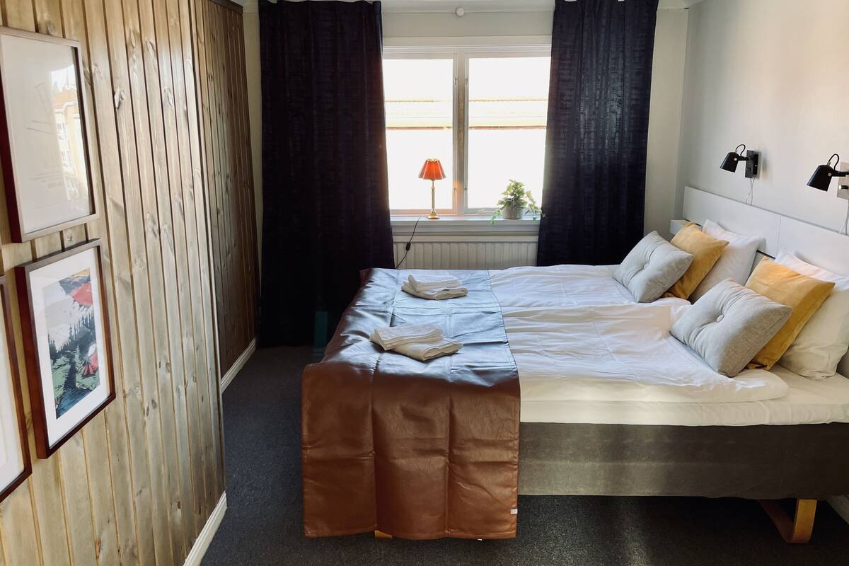 Quarto de hotel com duas camas, parede revestida de madeira, janela com cortinas, luminária e iluminação embutida na parede.