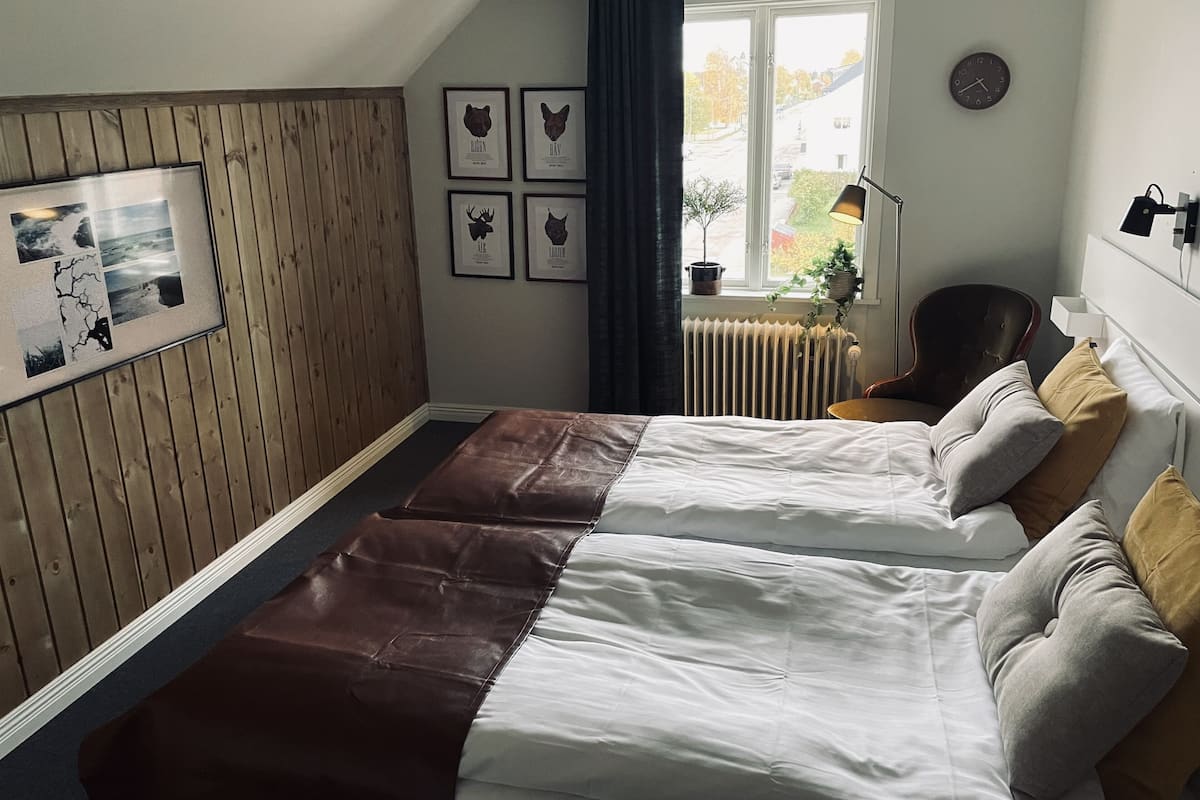 Quarto com duas camas, janela com vista, radiador e quadros na parede.