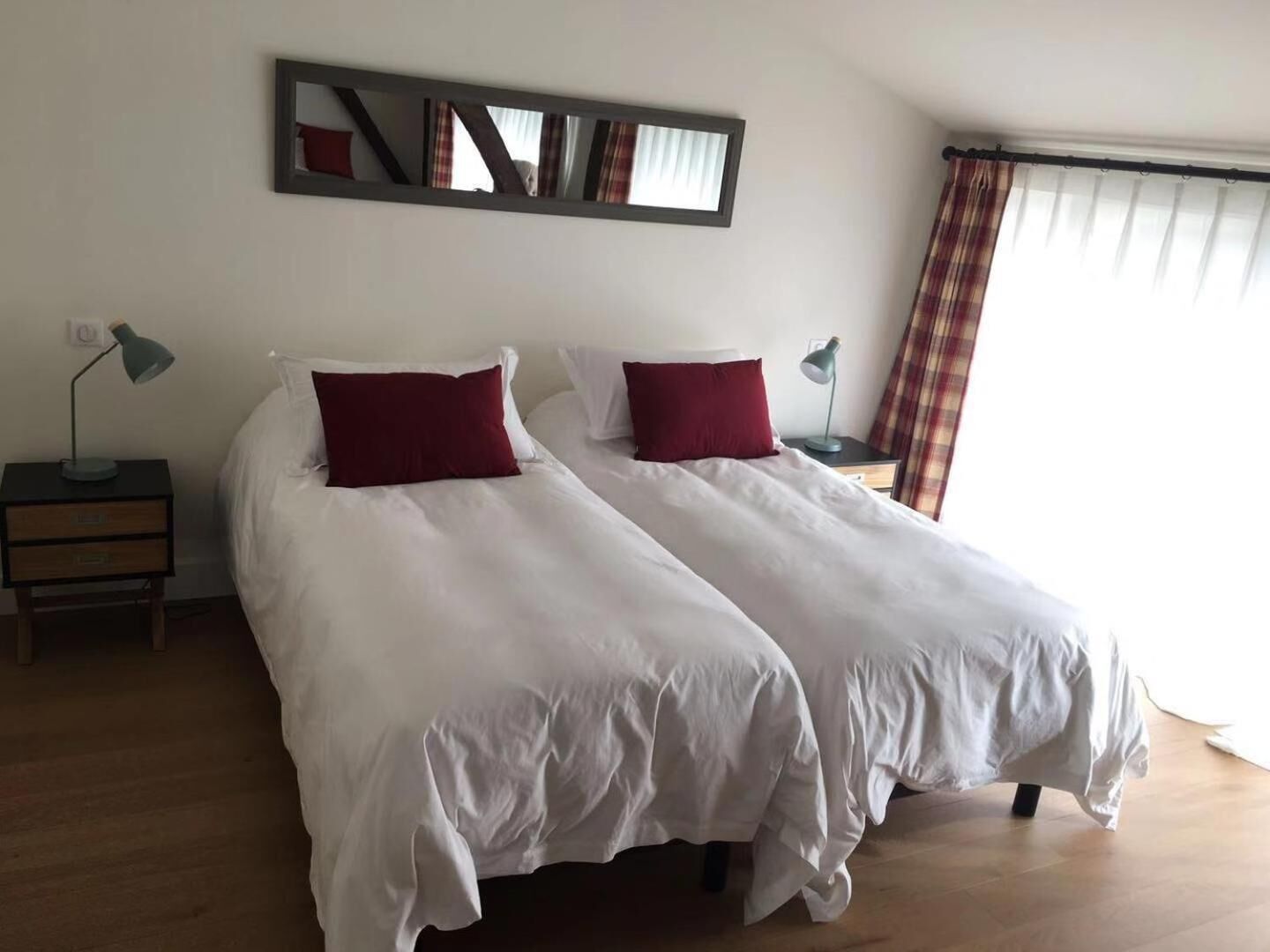 Uma cama de casal com roupa de cama branca e almofadas vermelhas, uma mesinha de cabeceira com abajur e um espelho na parede.
