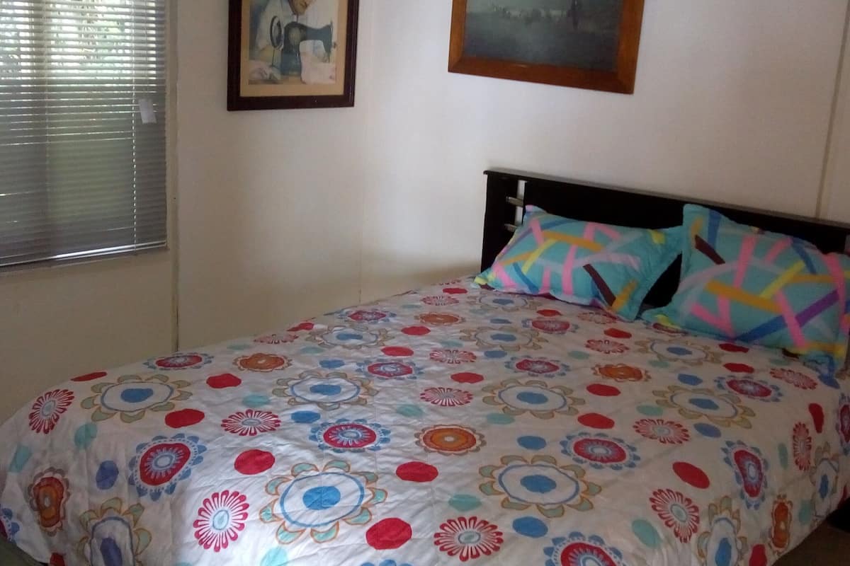Quarto com cama, roupa de cama floral colorida, janela com persianas e dois quadros na parede.