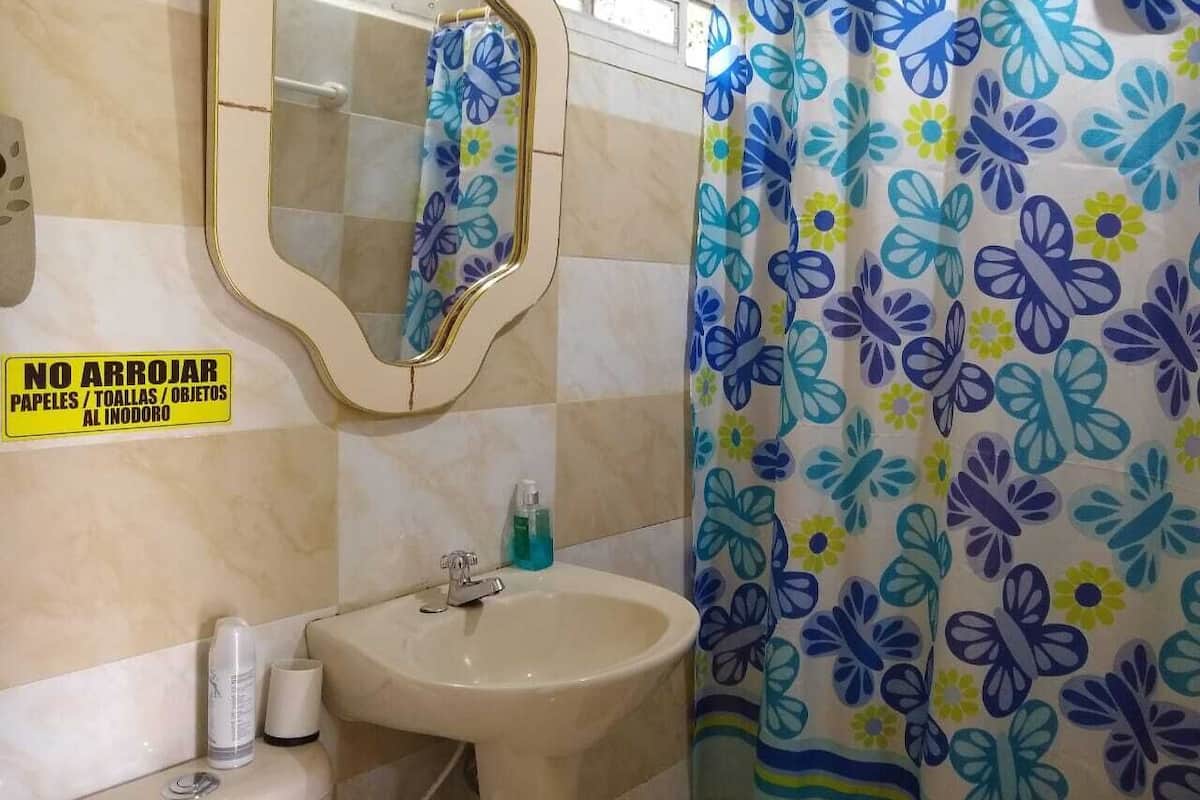 Casa de banho com lavatório, espelho e cortina de chuveiro com padrão de borboletas. Um aviso na parede diz "NÃO ATIRAR PAPÉIS/TOALHAS/OBJETOS NO SANITÁRIO.".