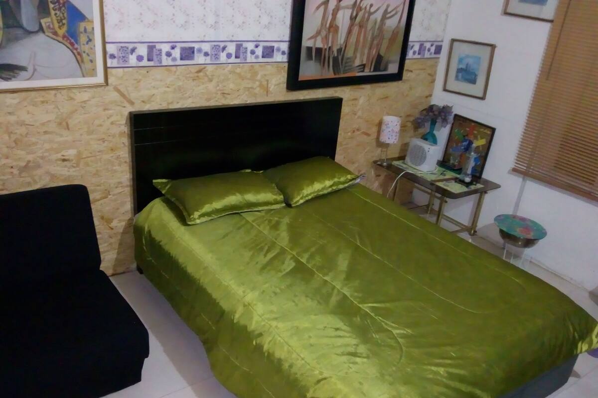 Quarto com cama, uma mesinha com um ventilador e janela com persianas.