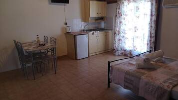 Quarto compacto com cama, pequeno espaço de cozinha, mesa de jantar com cadeiras e TV fixada na parede.