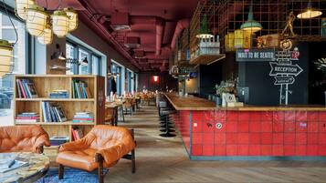 Uma área de bar com balcão vermelho, banquetas e um balcão de recepção. Há estantes de livros e poltronas de couro na área de estar.