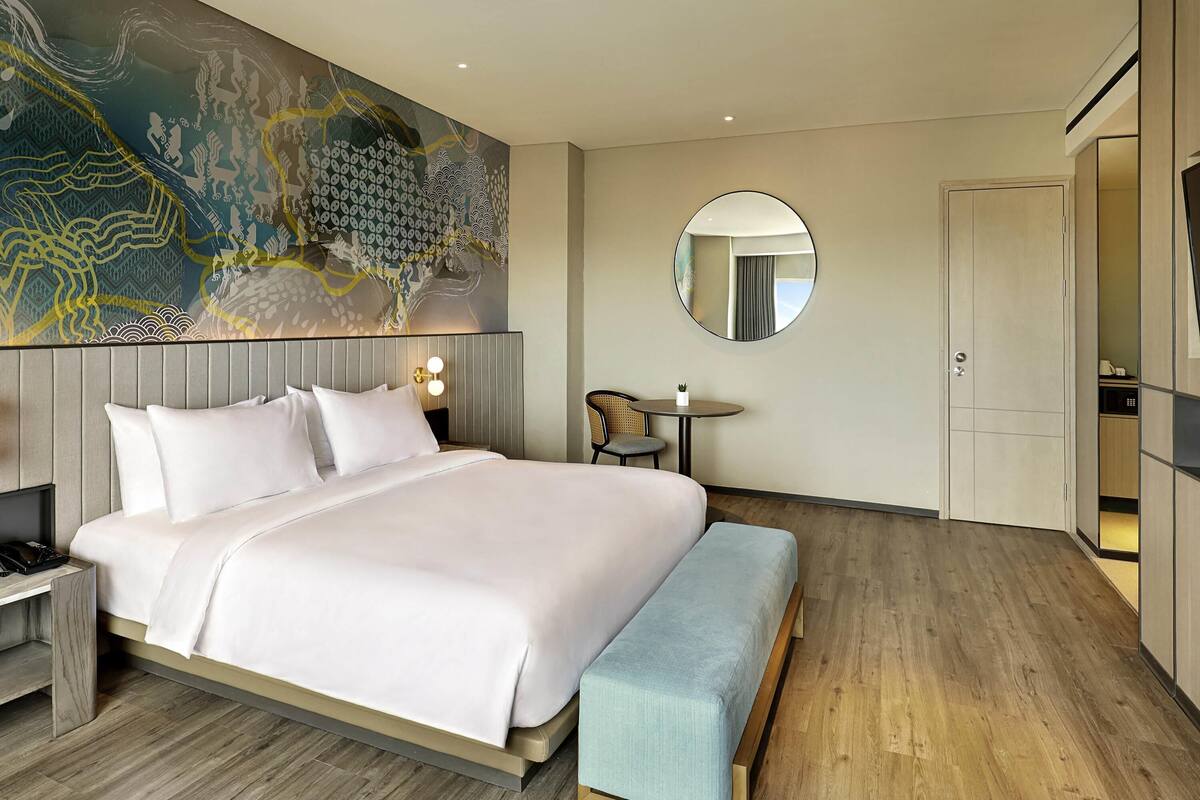 Quarto de hotel moderno com uma cama grande, mesas de cabeceira, um espelho redondo e uma obra de arte na parede.