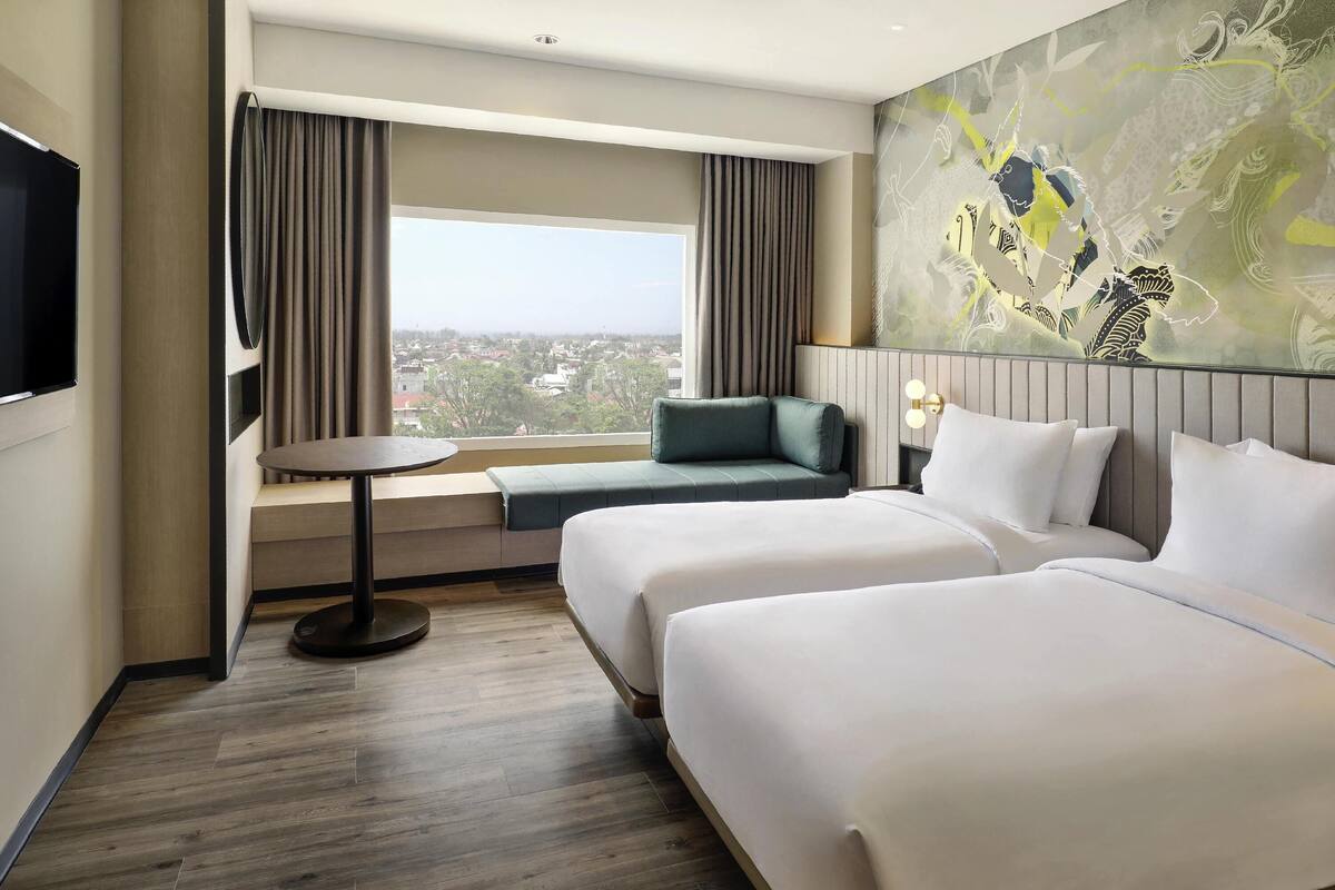 Quarto de hotel com duas camas, um sofá, uma mesinha e uma janela grande com cortinas.