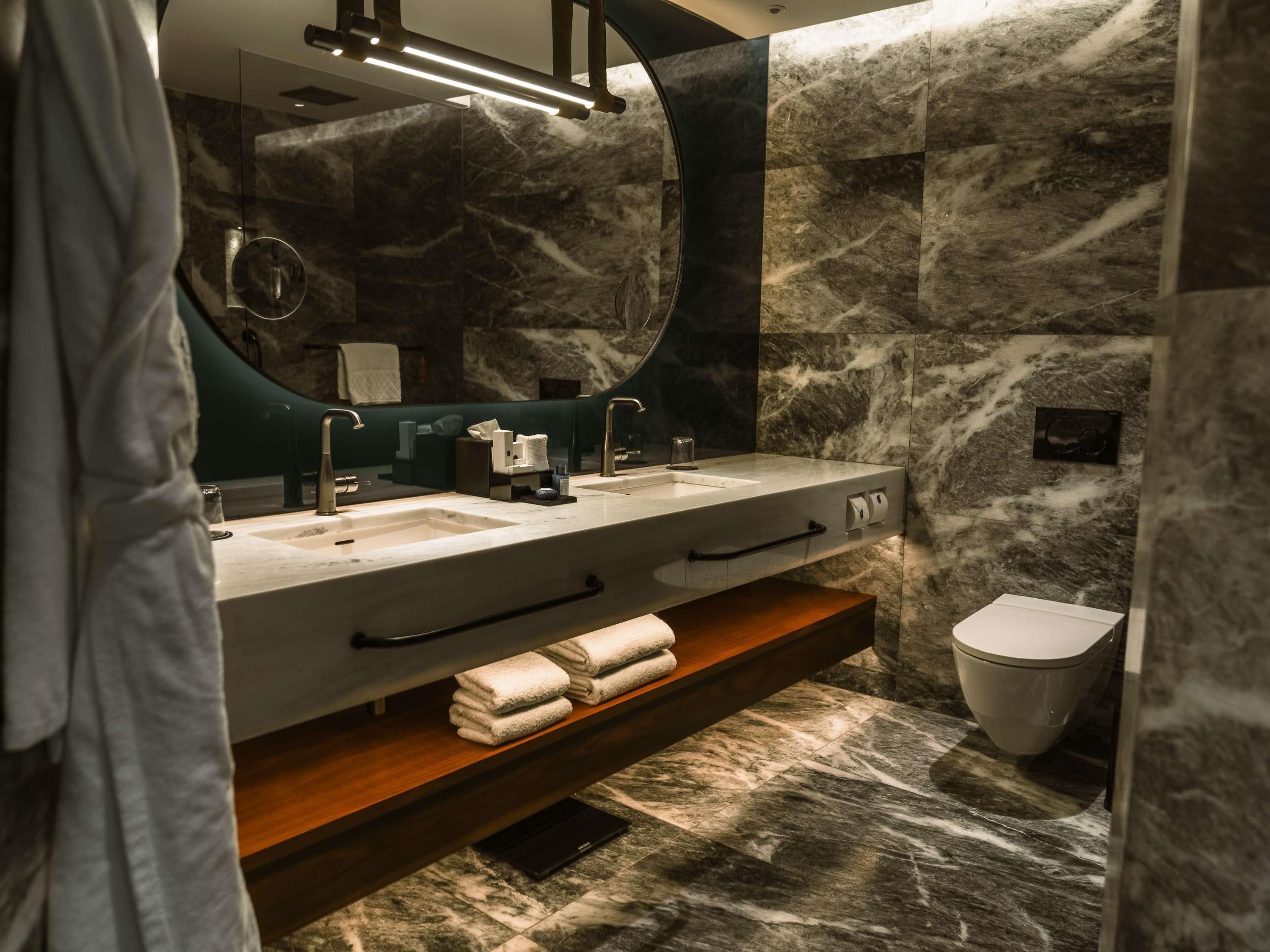 Um banheiro moderno com parede de mármore, um espelho grande, duas pias, um vaso sanitário e um móvel de banheiro em madeira.