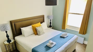 Cama bem arrumada com colcha azul, almofadas brancas e cabeceira marrom. Há duas luminárias de apoio com abajures estampados, uma pequena mesa com luminária e uma janela com cortinas.