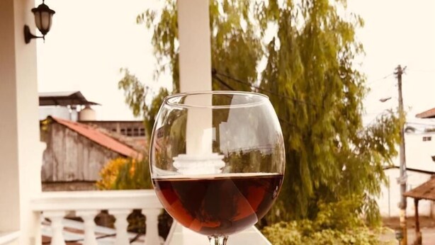 Um copo de vinho tinto sobre uma mesa branca, em frente a um edifício com guarda-corrimão branco e uma árvore ao fundo.