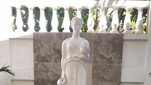 Estátua de mármore branco de uma mulher segurando um objeto pequeno, sobre um pedestal com base de mármore, rodeada por plantas em vasos.