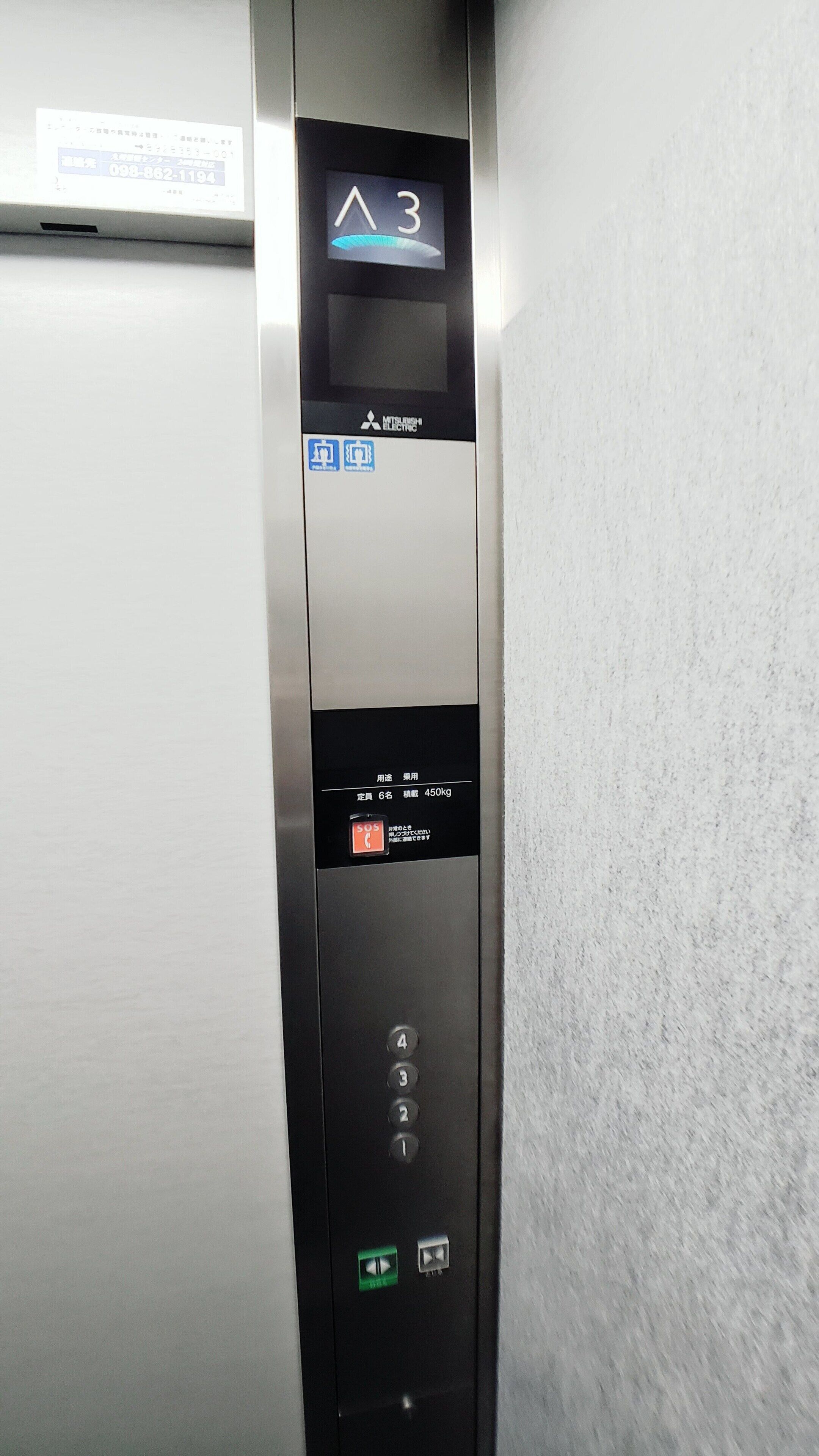 Painel de comando de elevador com logotipo Mitsubishi, botão de emergência e botões de seleção de andar.