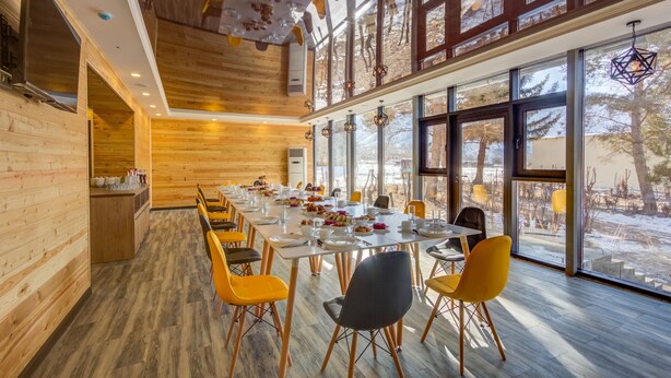 Uma sala de jantar espaçosa com uma mesa comprida posta para uma refeição, grandes janelas com vista para o exterior e painéis de madeira nas paredes.