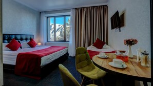 Quarto de hotel com duas camas, televisão, mesa de jantar com jogo de chá e janela com vista para montanhas nevadas.