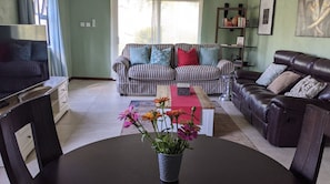 Uma sala de estar com um sofá de padrão listrado, um sofá de couro, uma mesa de centro com um vaso de flores e uma televisão sobre um suporte.