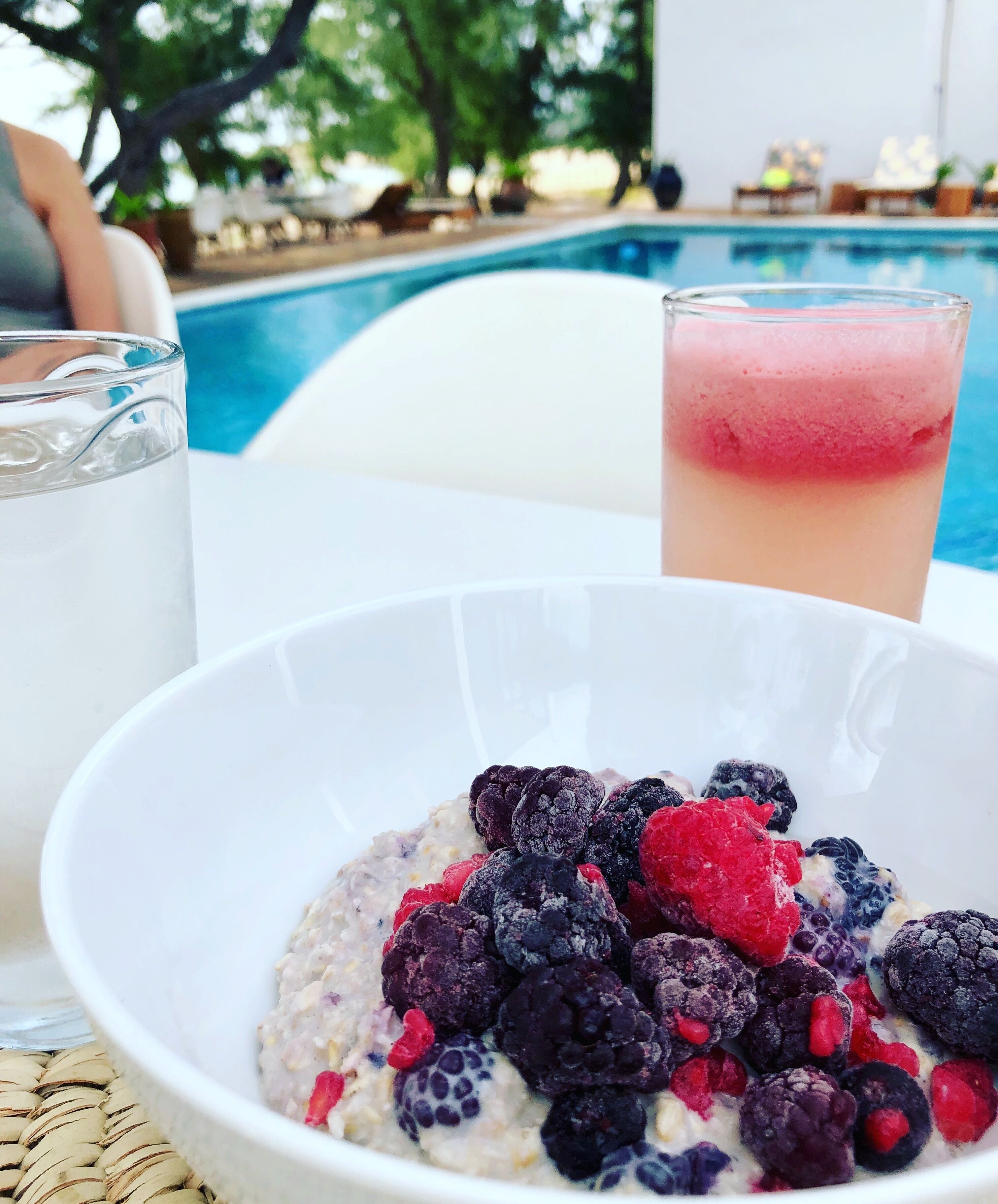 Uma tigela de aveia com frutas vermelhas, um copo de água e um smoothie à beira da piscina.