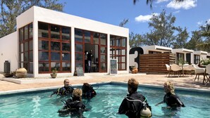 Uma área de piscina com água azul cristalina, rodeada por um edifício moderno com grandes janelas e fachada branca.