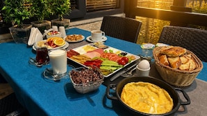 Mesa de café da manhã com diversos alimentos, uma xícara de café e vista ensolarada para o exterior.
