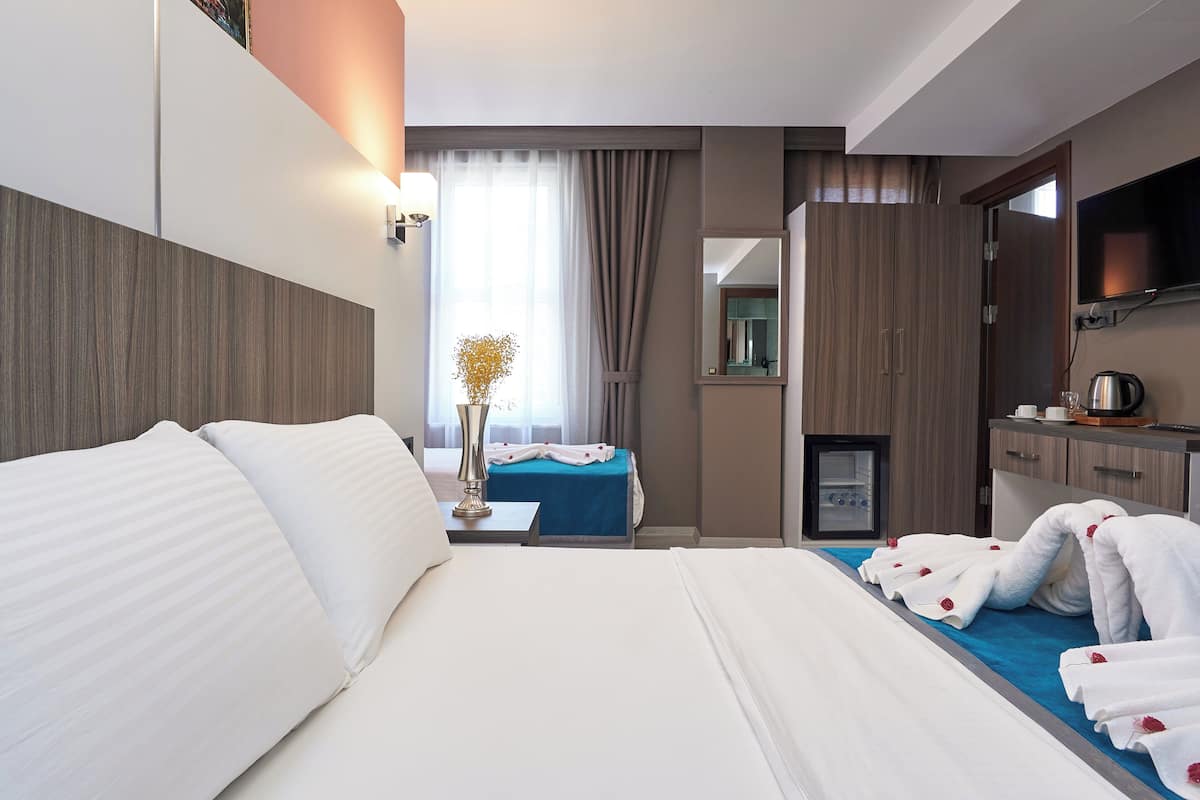 Quarto de hotel com uma cama grande, uma mesa com luminária, minibar, televisão e banheiro visível através de uma porta aberta.