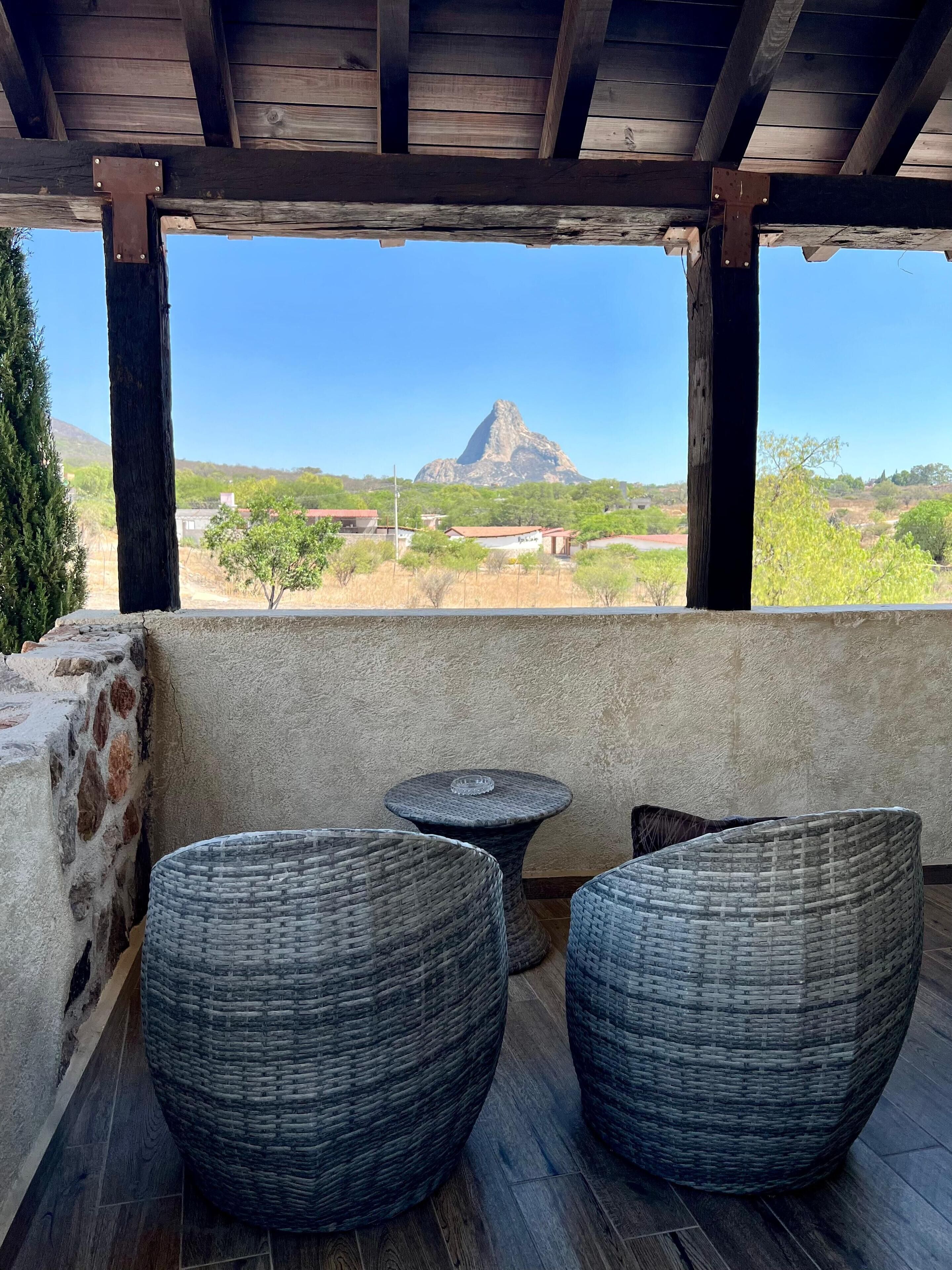 Uma área de estar coberta ao ar livre, com móveis de vime e vista para uma montanha.