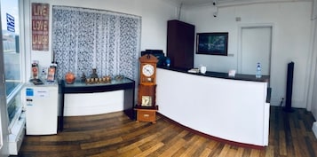 Quarto com piso de madeira, balcão de recepção branco, um relógio e uma janela com cortinas florais.