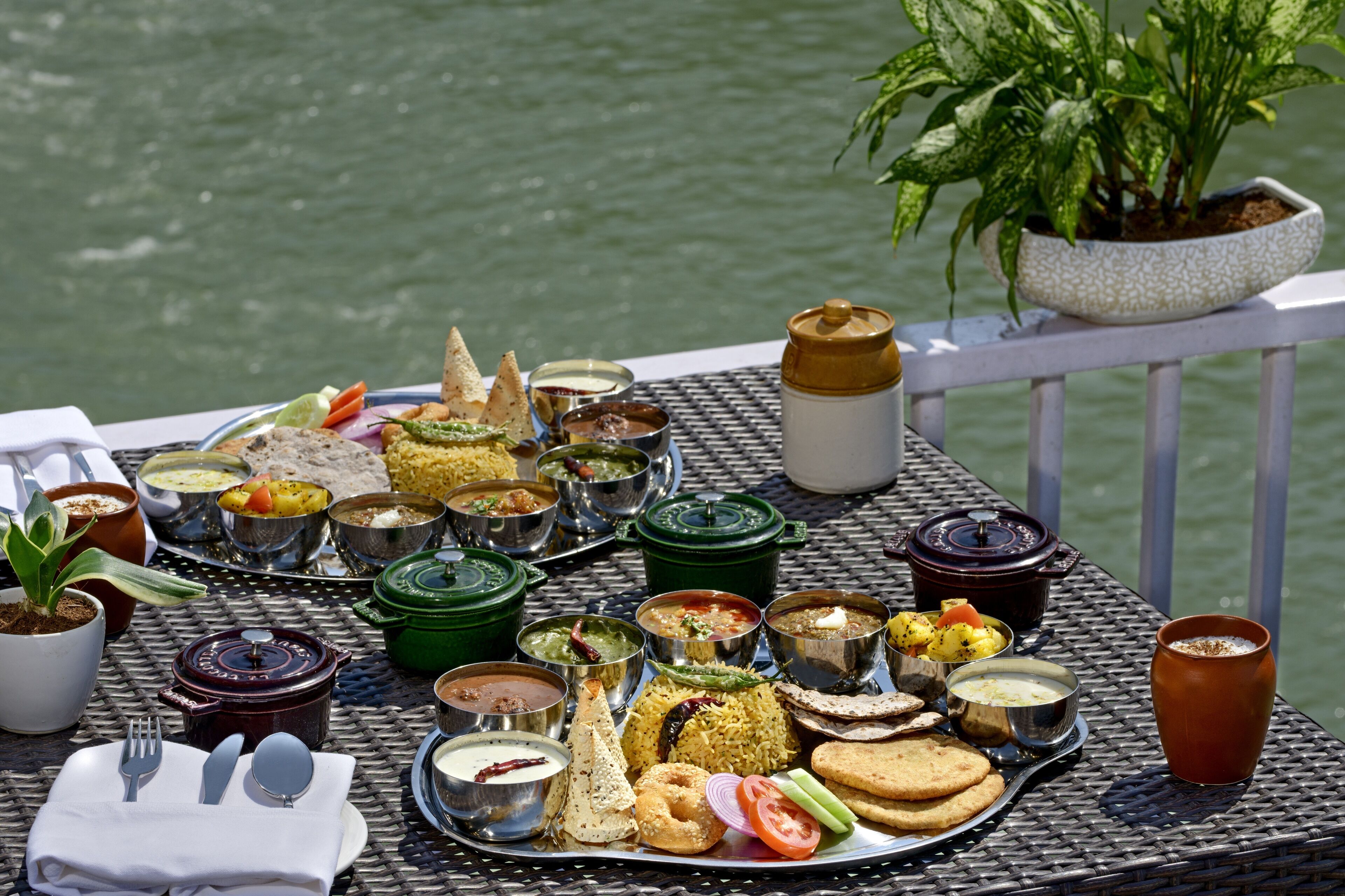 Mesa posta com diversos pratos e bebidas, possivelmente para uma refeição, em uma varanda com vista para a água.