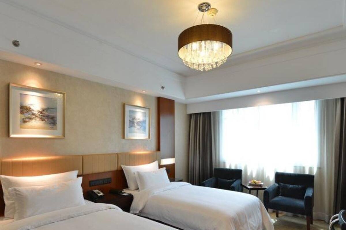 Quarto de hotel com duas camas, um candelabro, uma escrivaninha com cadeira e uma janela grande com cortinas.