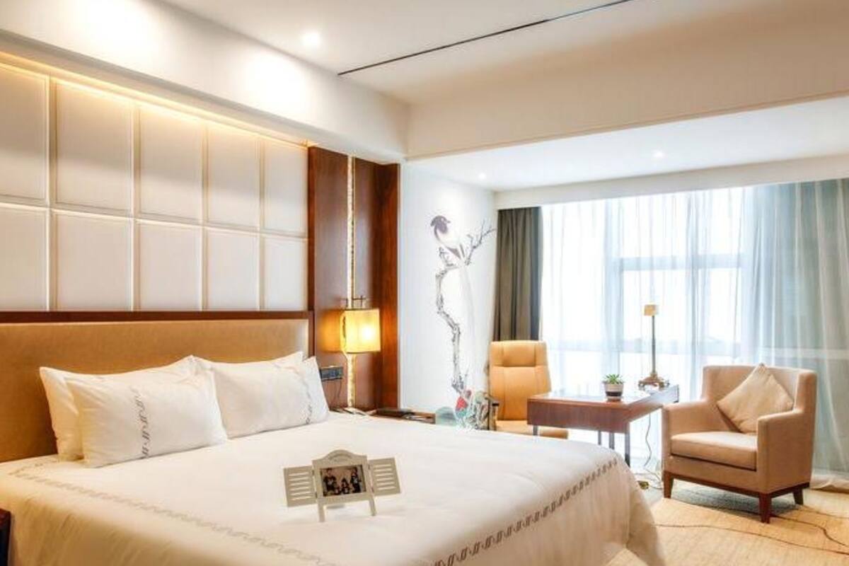 Quarto de hotel com cama grande, uma cadeira, escrivaninha e janela com cortinas.