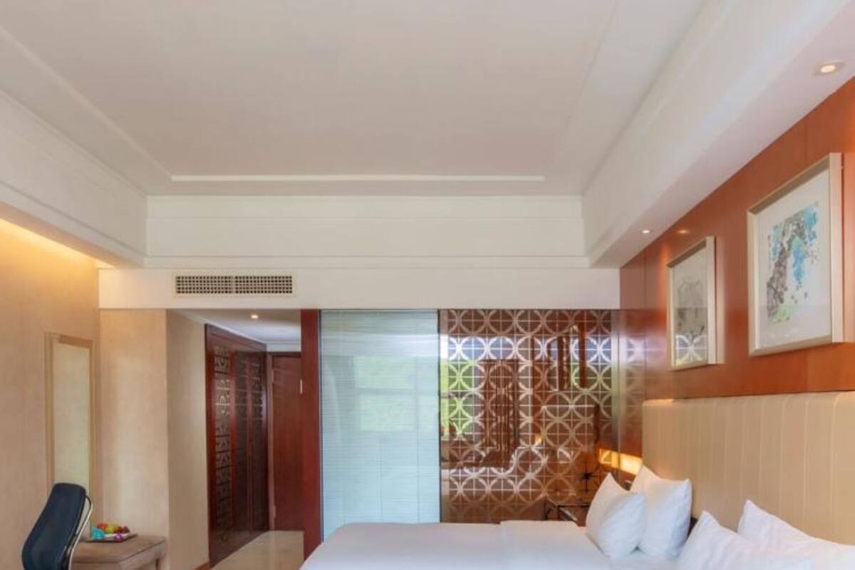 Quarto de hotel com uma cama grande, uma escrivaninha e uma cadeira. Há uma janela com persianas e uma porta que dá para outro quarto.