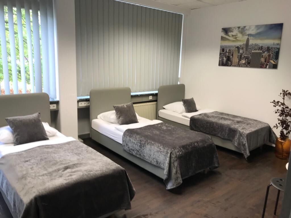 Quarto de hotel com três camas de solteiro, um quadro de uma paisagem urbana na parede e uma janela com persianas.