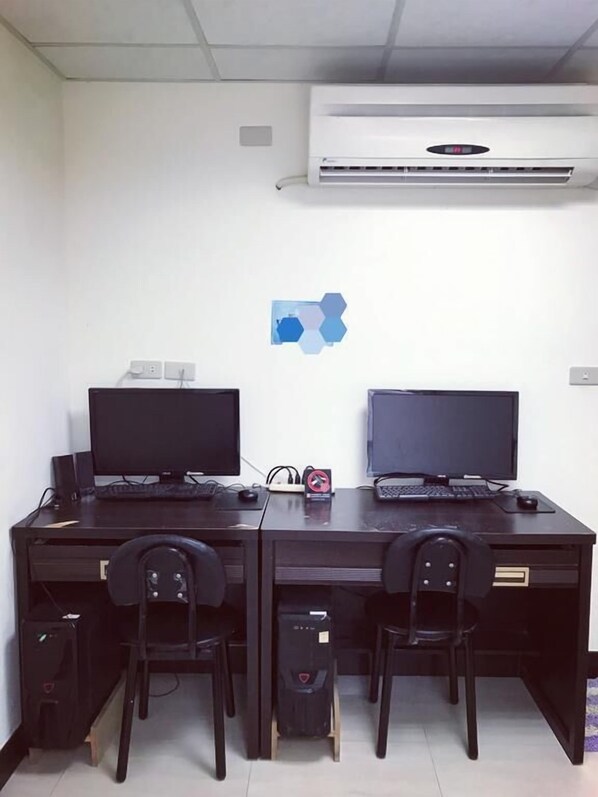 Um espaço de trabalho com duas mesas, cada uma com monitor de computador, teclado e rato. Há duas cadeiras, uma em frente a cada mesa. Um aparelho de ar condicionado está montado na parede acima das mesas.