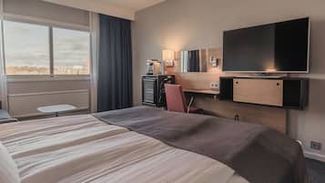 Quarto de hotel com uma cama grande, uma escrivaninha com cadeira, televisão de tela plana e vista para a cidade pela janela.