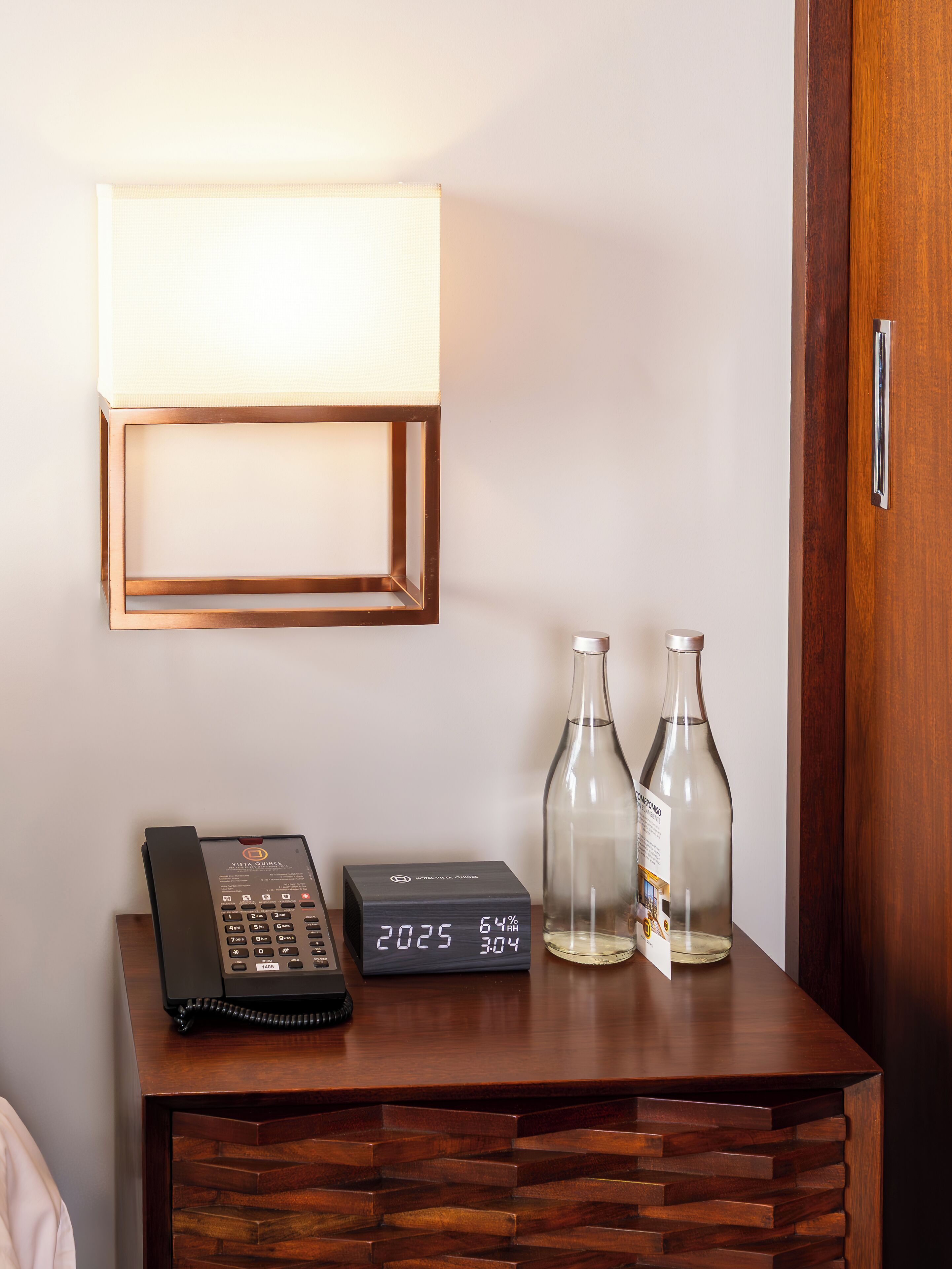 Uma mesa de cabeceira de madeira com um telefone preto, um relógio digital mostrando 2025 64% AM e duas garrafas de vidro vazias.