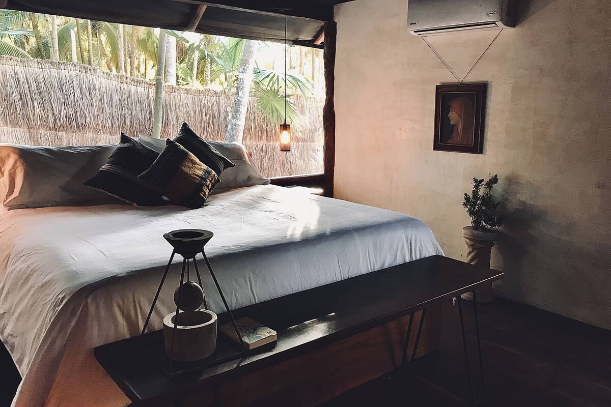 Quarto com cama, almofadas, mesa de cabeceira com abajur, planta em vaso e ar condicionado.
